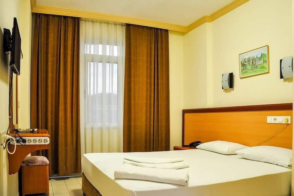 Quarto de hotel com cama, cortinas, televisão, mesa de cabeceira e um quadro pendurado na parede.