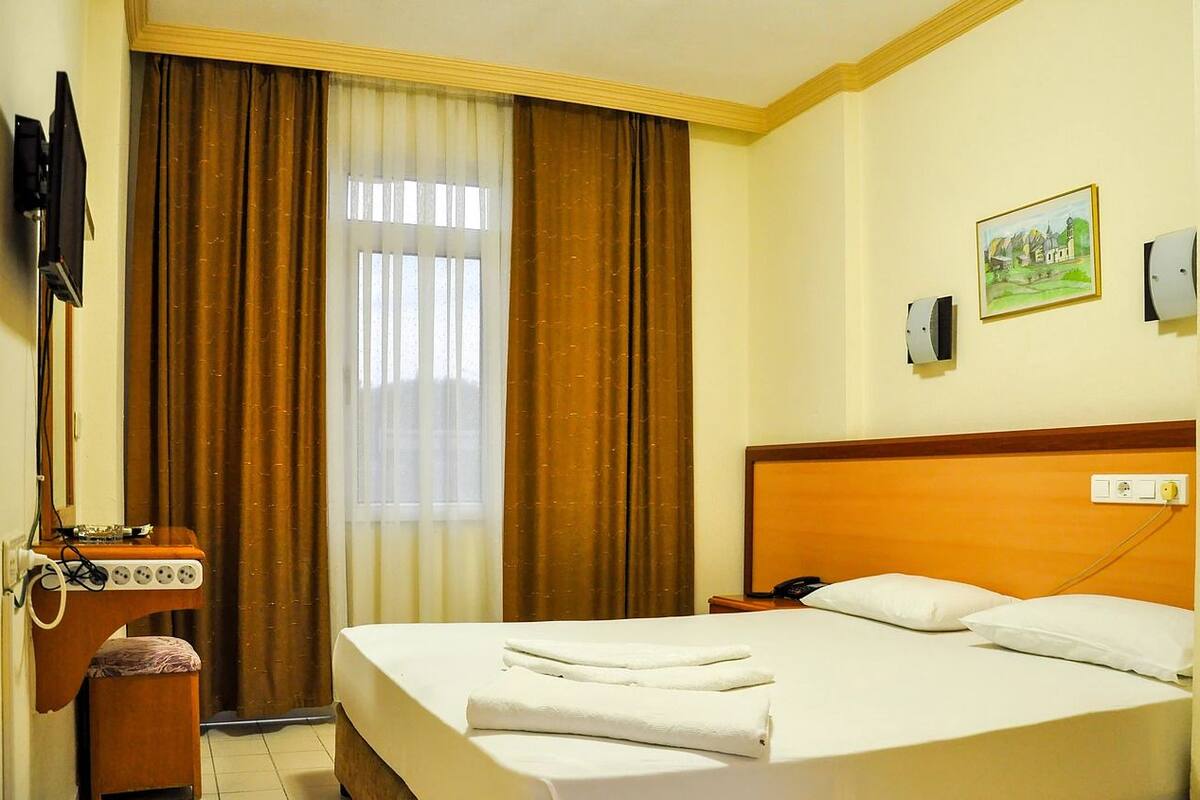 Quarto de hotel com cama, escrivaninha com telefone, televisão e um quadro na parede.