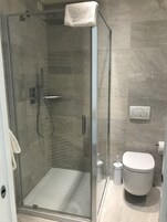 Casa de banho moderna com chuveiro em cabine de vidro, sanita e toalha pendurada na porta.