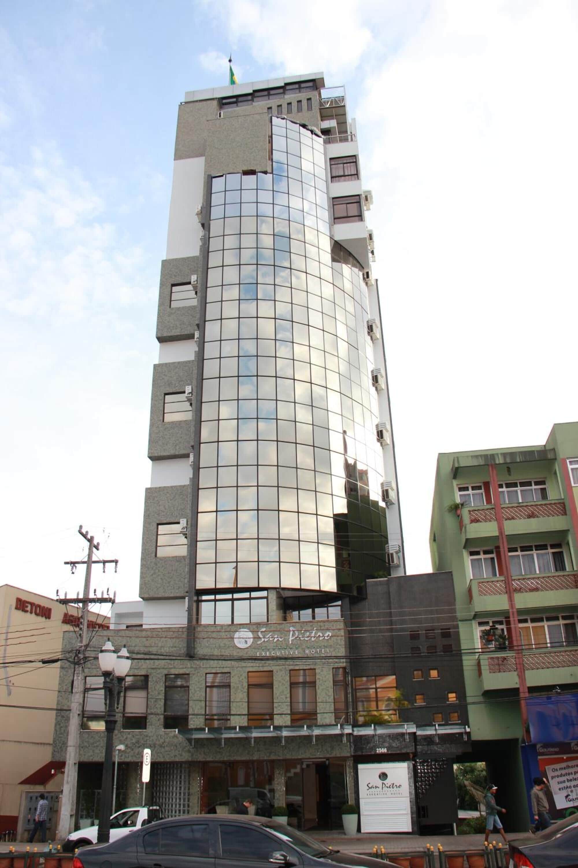 Um edifício alto com fachada de vidro refletante, várias varandas e uma placa com o nome 'San Pietro Executive Hotel' na base.