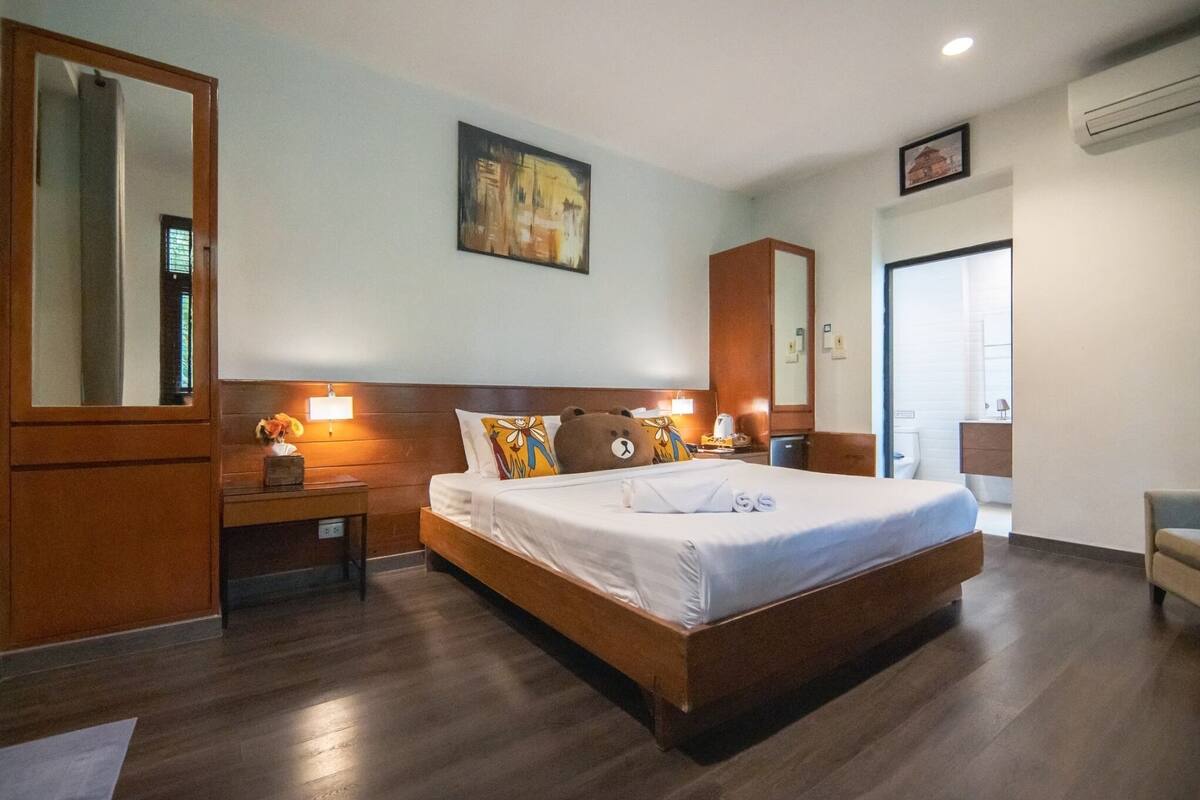 Quarto de hotel com uma cama grande, cabeceira de madeira, mesas de cabeceira com abajures, um espelho, um quadro na parede e uma porta que dá para o banheiro.