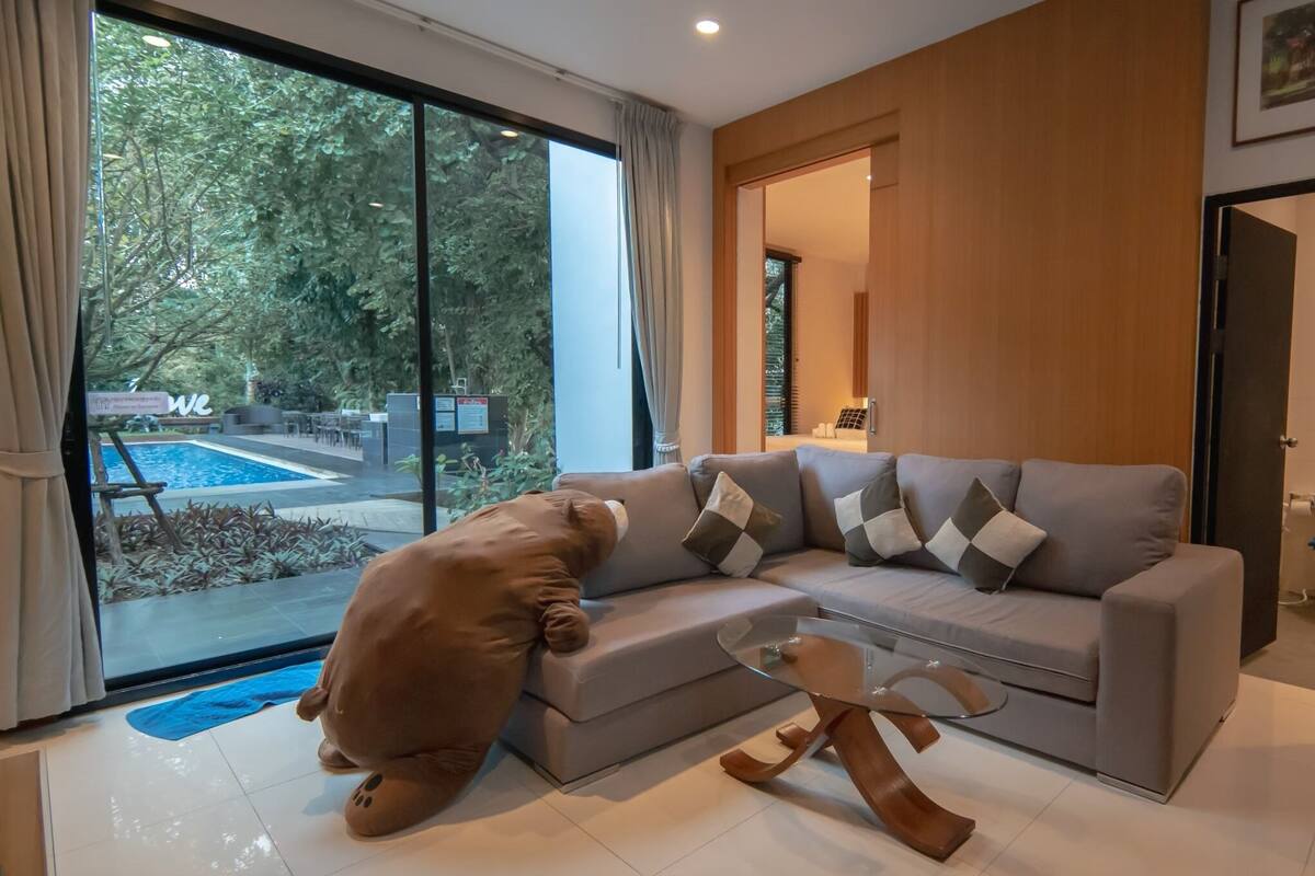 Uma sala de estar moderna com um sofá cinzento, uma mesa de centro de vidro e uma janela ampla com vista para uma piscina e vegetação.