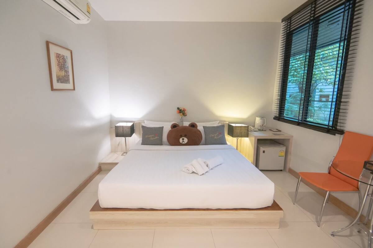 Quarto de hotel bem arrumado, com uma cama grande, mesa de cabeceira com abajur, escrivaninha pequena com cadeira e janela com persianas.