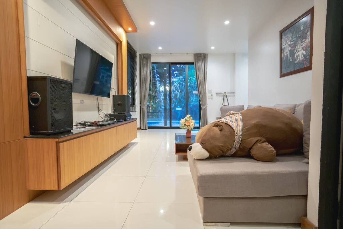 Sala de estar moderna com televisão de tela plana, mobiliário de madeira, um sofá com um ursinho de pelúcia e vista para árvores pela porta de vidro.