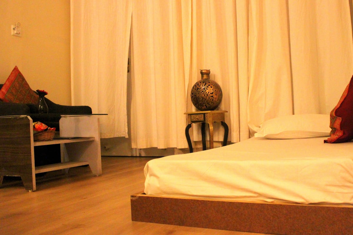 Quarto com cama, mesa de cabeceira, cadeira, pequena mesa e um vaso.