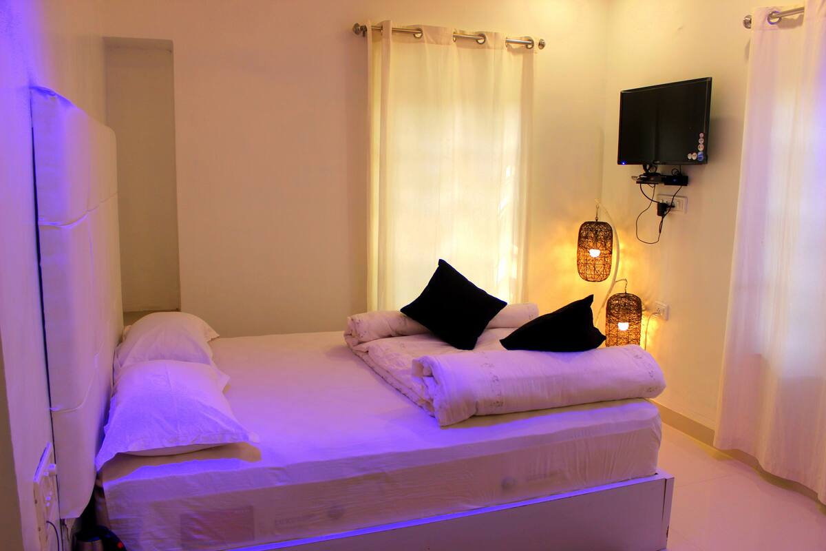Cama bem arrumada com travesseiros, televisão montada na parede e luminária fixada na parede em um quarto com cortina.