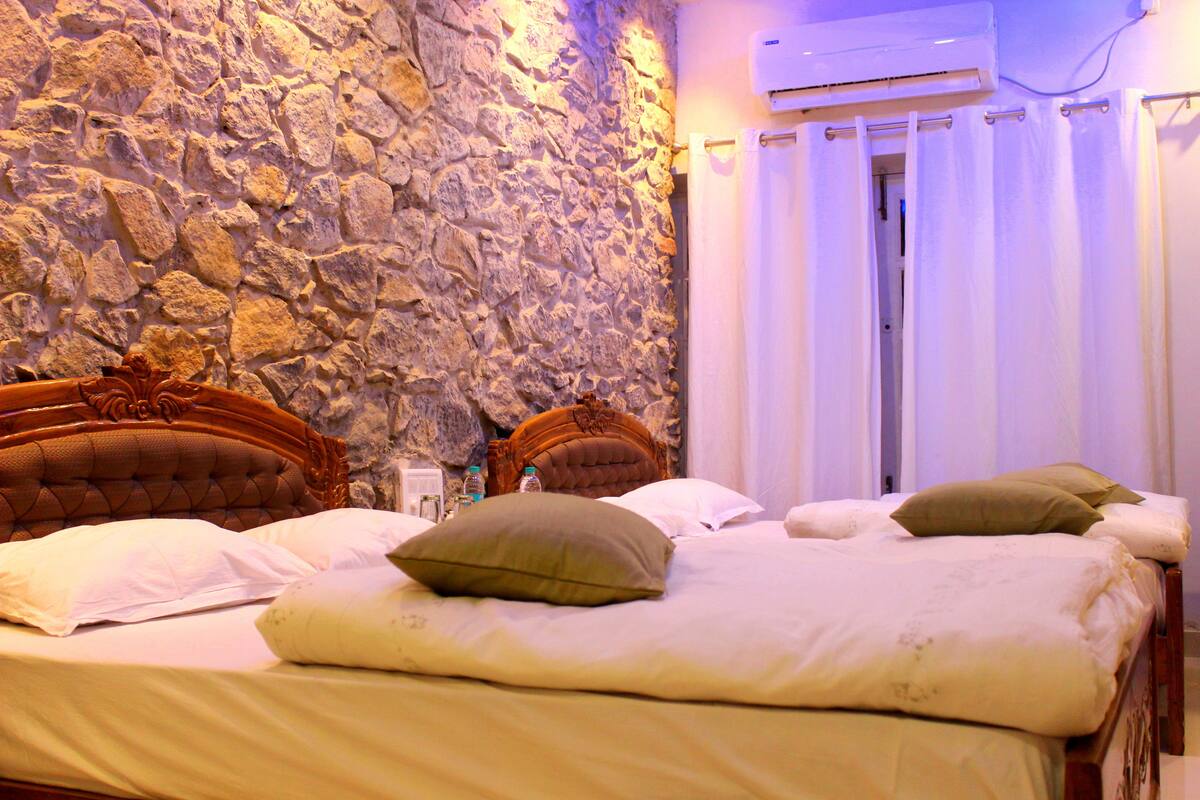 Quarto de hotel com parede de pedra, duas cabeceiras de madeira e uma cama com roupa de cama branca e almofadas verdes.