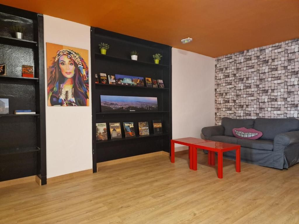 Uma sala de estar com televisão de tela plana, uma estante com livros e objetos decorativos, um sofá cinza com almofada divertida e uma mesa de centro vermelha.