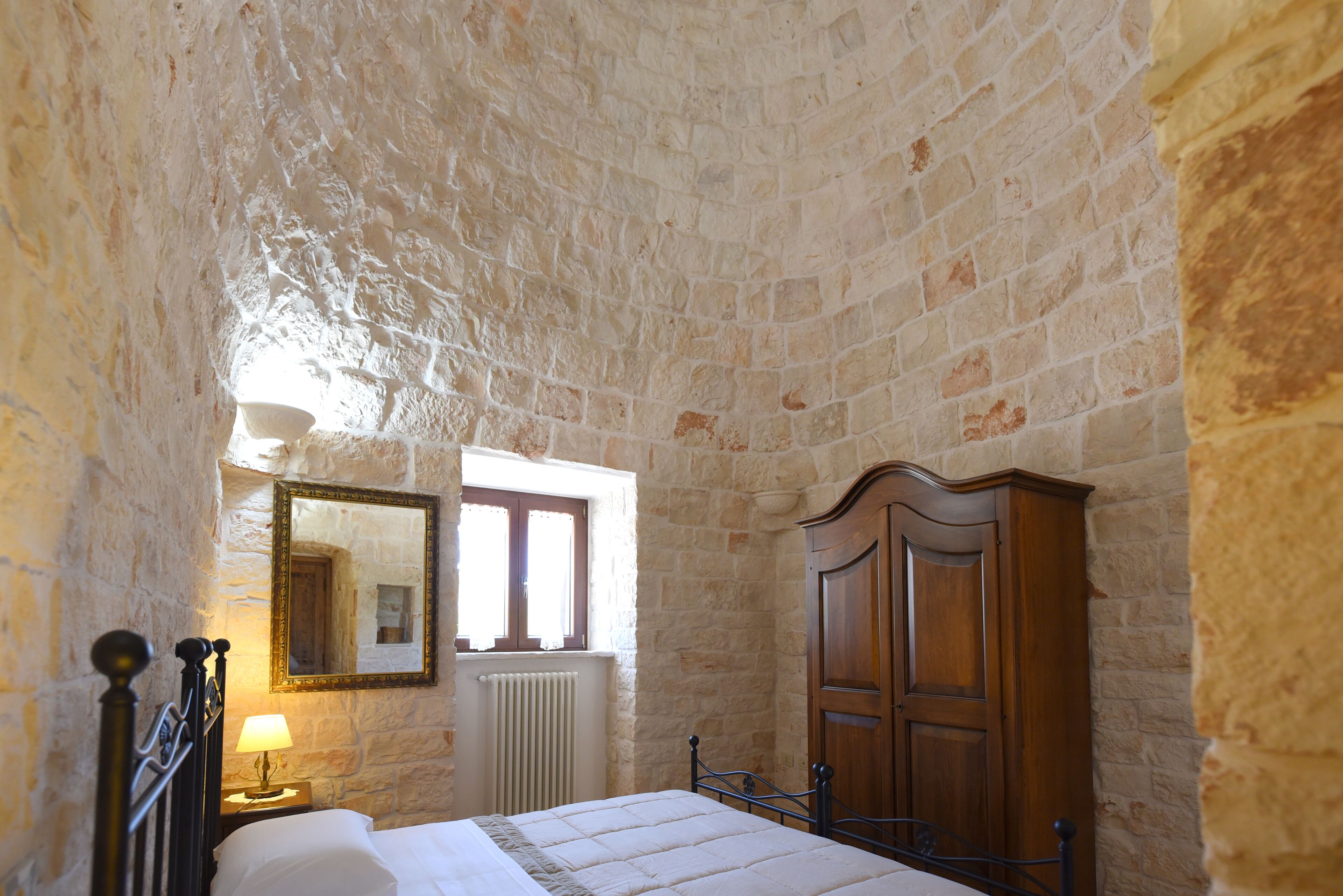 Quarto com paredes de pedra, uma cama de madeira, uma mesa de cabeceira com abajur, um guarda-roupa de madeira e uma janela com espelho.