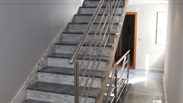 Uma escadaria moderna com degraus de mármore e guarda-corpos de metal, que leva a uma porta no topo.