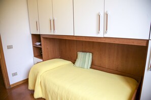 Quarto pequeno com uma cama de solteiro, cabeceira de madeira, colcha amarela e armários brancos acima.