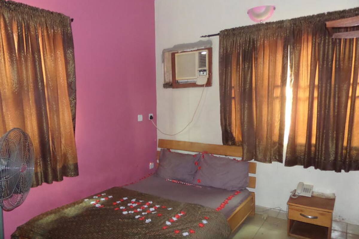 Quarto com cama, mesa de cabeceira, ventilador, aquecedor e cortinas.