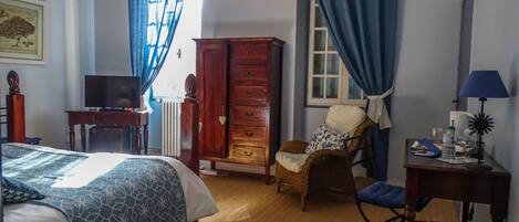 Quarto de hotel com cama, escrivaninha, cadeira, cômoda, abajur e janela com cortinas.