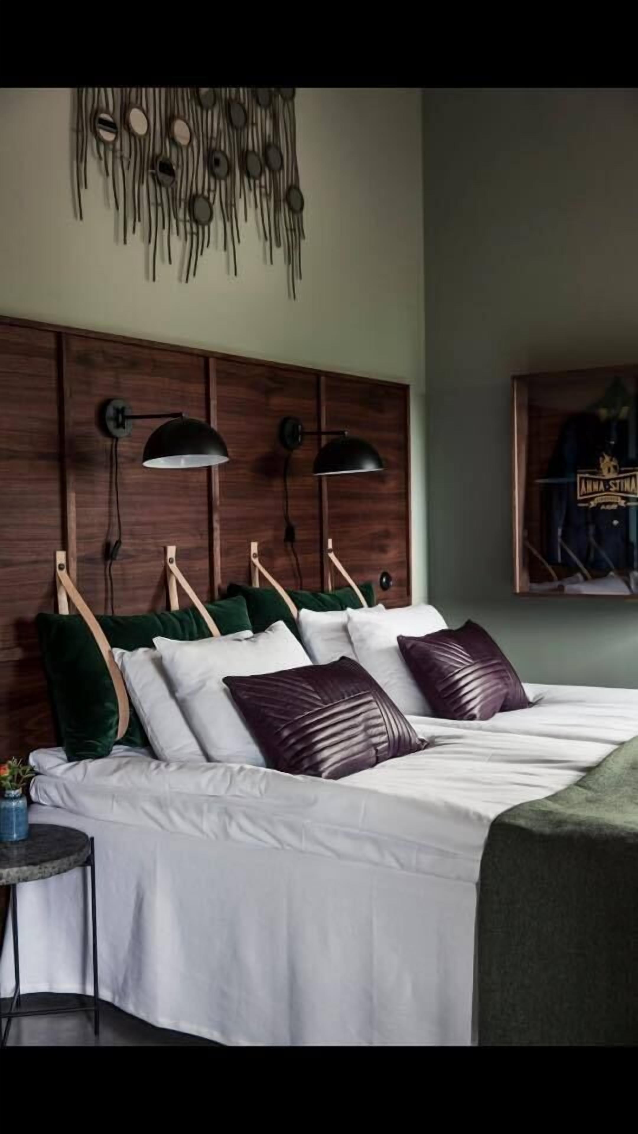 Quarto com cabeceira de madeira, luminária pendente e cama com roupa de cama branca e várias almofadas.