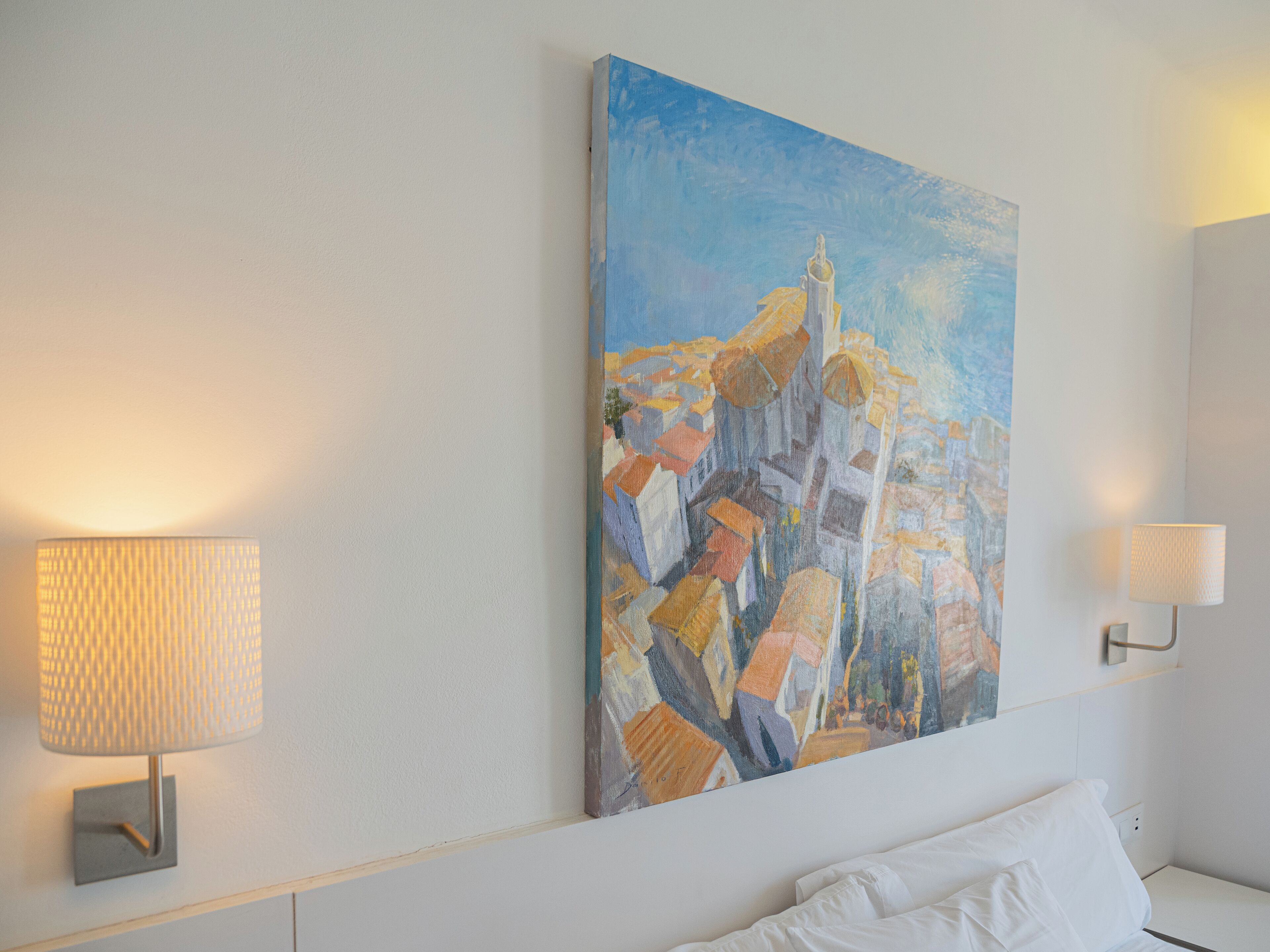 Quarto de hotel com uma grande pintura de paisagem urbana na parede, uma cama com lençóis brancos e duas luminárias fixadas na parede.