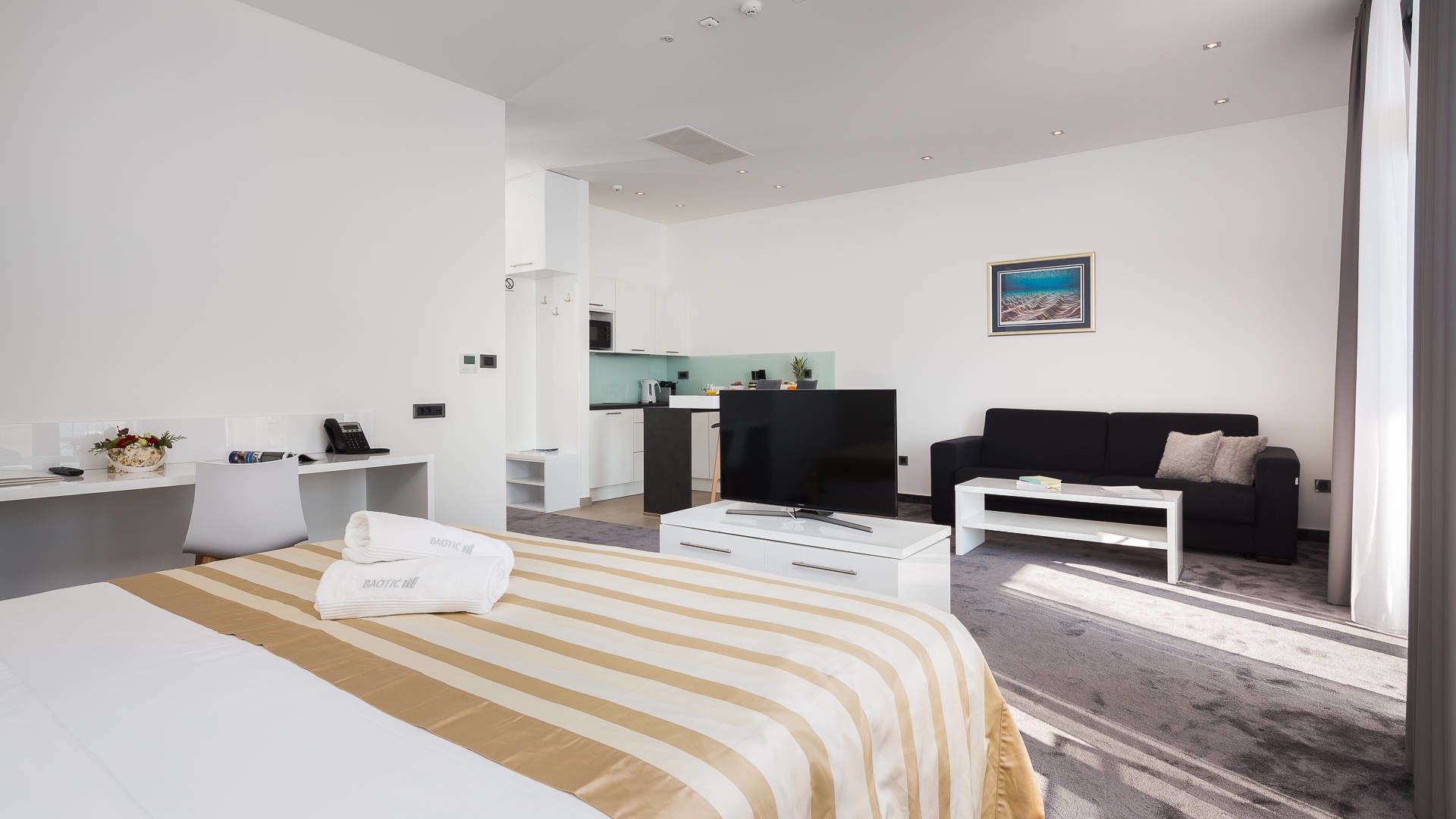 Quarto de hotel moderno com uma cama grande, uma escrivaninha, um sofá preto e uma área de cozinha ao fundo.