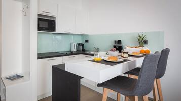 Uma cozinha compacta com armários brancos, bancada preta e uma área de refeições com mesa branca e duas cadeiras cinzentas.