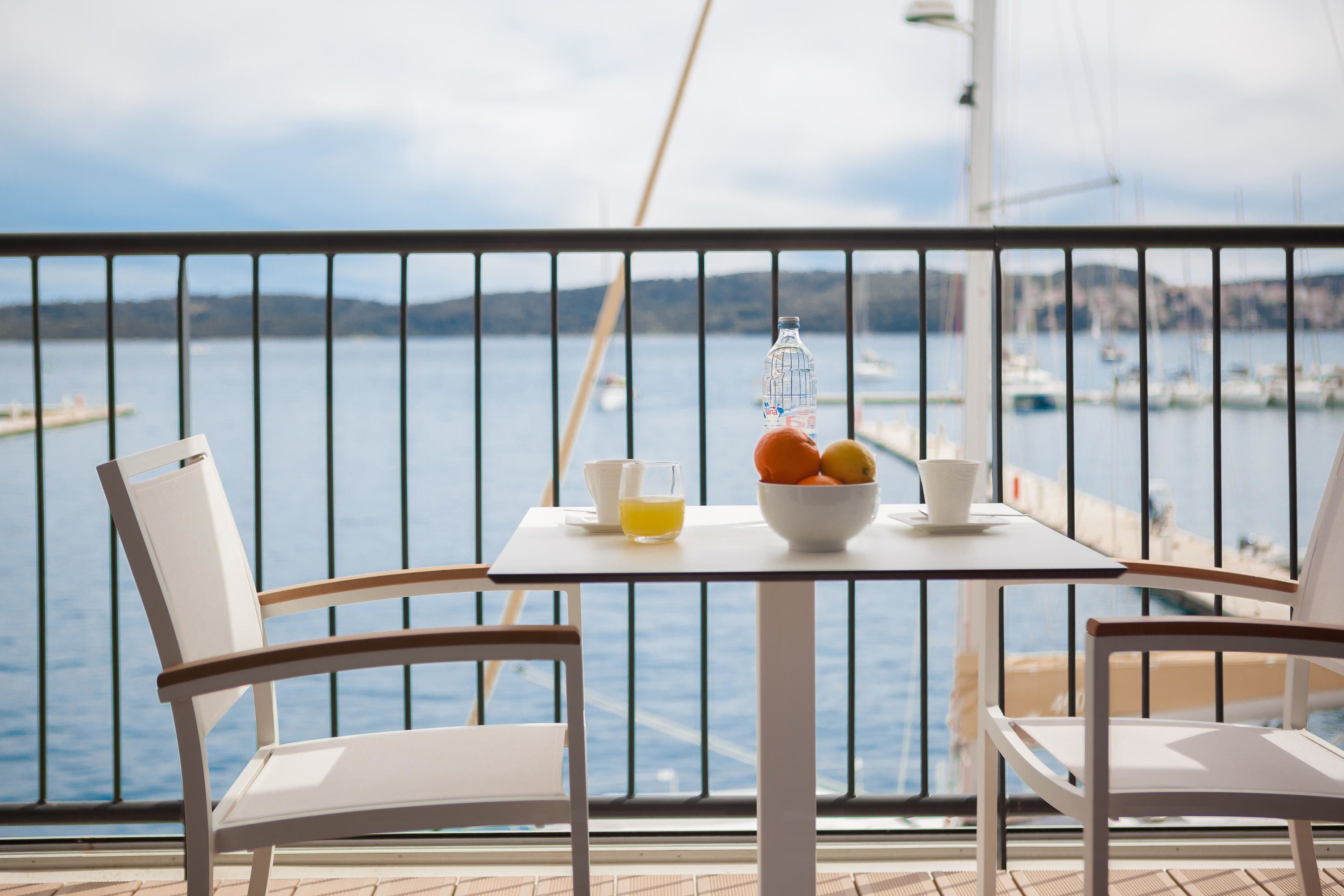 Varanda com mesa posta para dois, exibindo uma tigela de frutas, um copo de suco e uma garrafa de água. A vista inclui um porto com barcos e uma linha de costa ao longe.