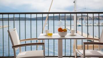 Varanda com mesa posta para dois, exibindo uma tigela de frutas, um copo de suco e uma garrafa de água. A vista inclui um porto com barcos e uma linha de costa ao longe.
