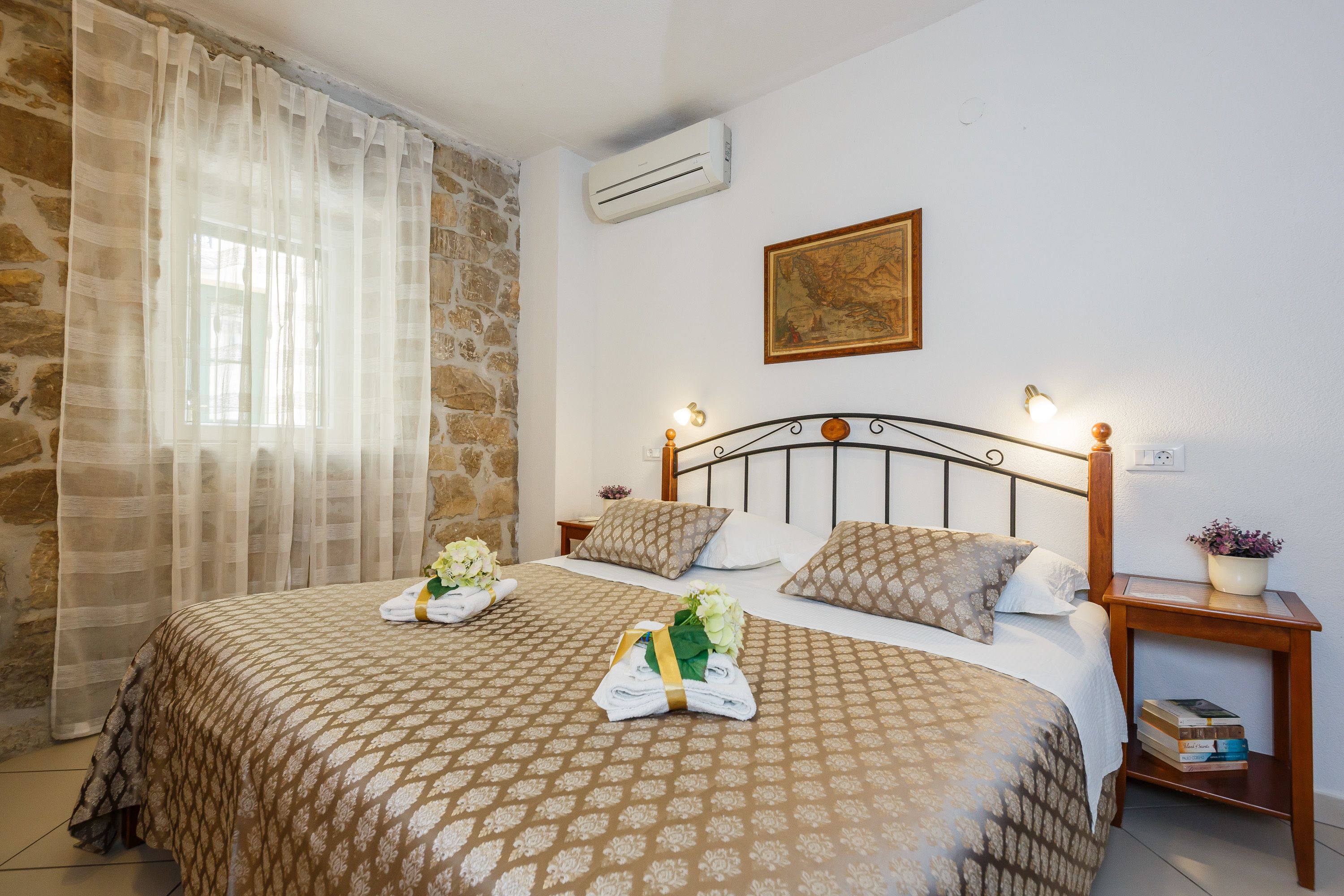 Uma cama de casal com colcha estampada, duas almofadas e um buquê de flores. Há uma mesa de cabeceira com abajur e uma pequena planta. O quarto tem paredes de pedra e um ar condicionado na parede.