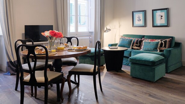 Uma sala de jantar com uma mesa redonda, cadeiras e um vaso de flores. Há um sofá verde com almofadas estampadas e um abajur.