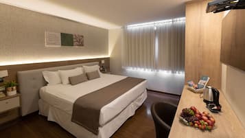Quarto de hotel com uma cama grande, mesa de cabeceira com abajur, pequena escrivaninha com chaleira e uma cesta de frutas.