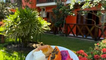 Uma mão segura um prato com arroz branco, uma mamão e uma flor rosa, com um jardim e um edifício ao fundo.