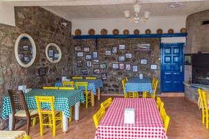 Uma área de refeições rústica com paredes de pedra, várias mesas com toalhas quadriculadas e cadeiras amarelas.