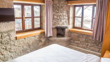 Quarto com lareira de pedra, janelas de madeira com cortinas, uma cama e televisão de tela plana na parede.