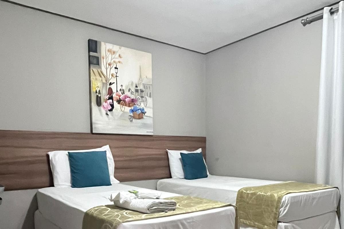 Quarto de hotel com duas camas, cabeceira, um quadro e janela com cortinas.