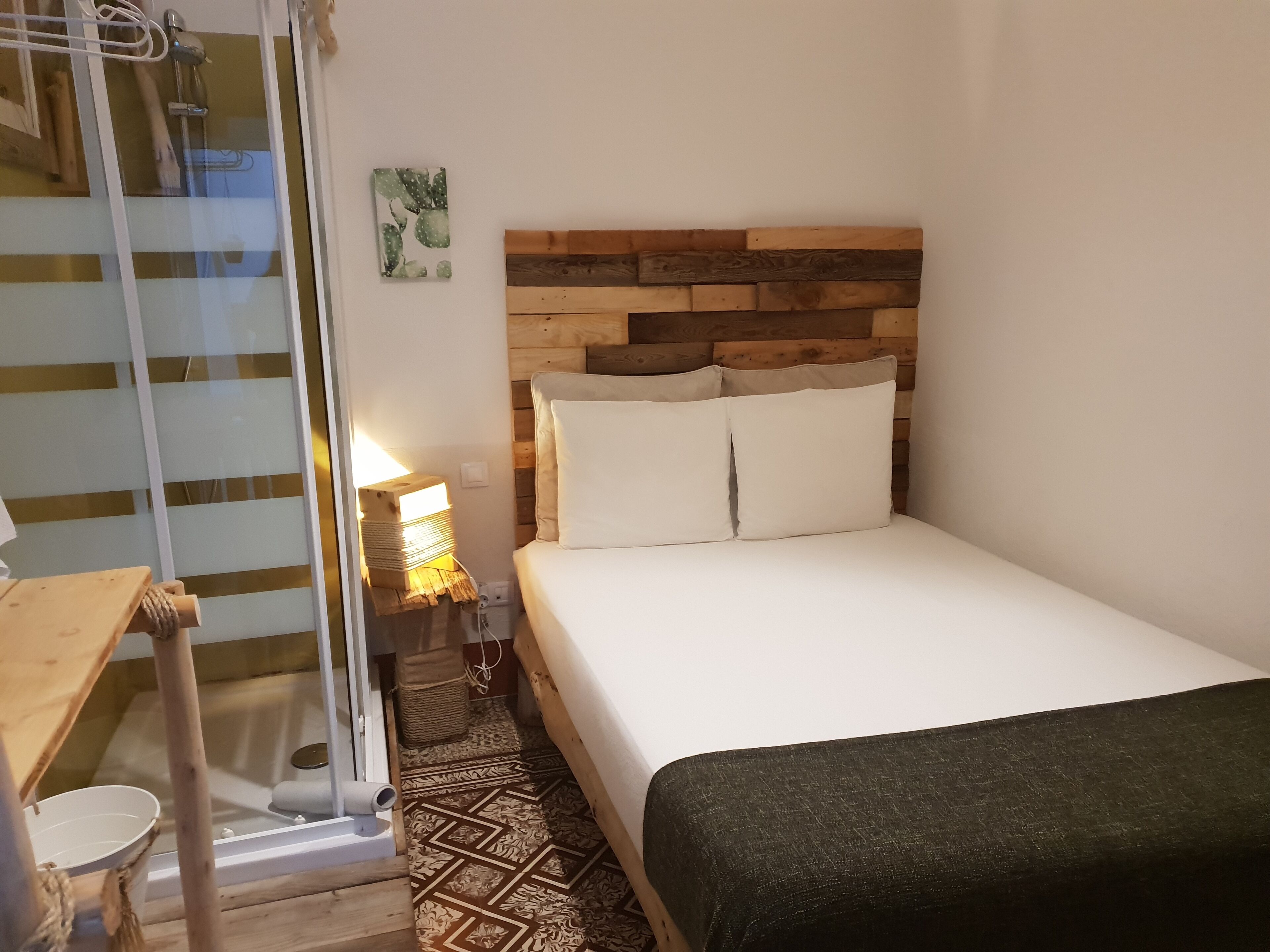 Um quarto pequeno e acolhedor com cabeceira de madeira, cama com roupa de cama branca, uma mesa pequena com abajur e um espaço com chuveiro.