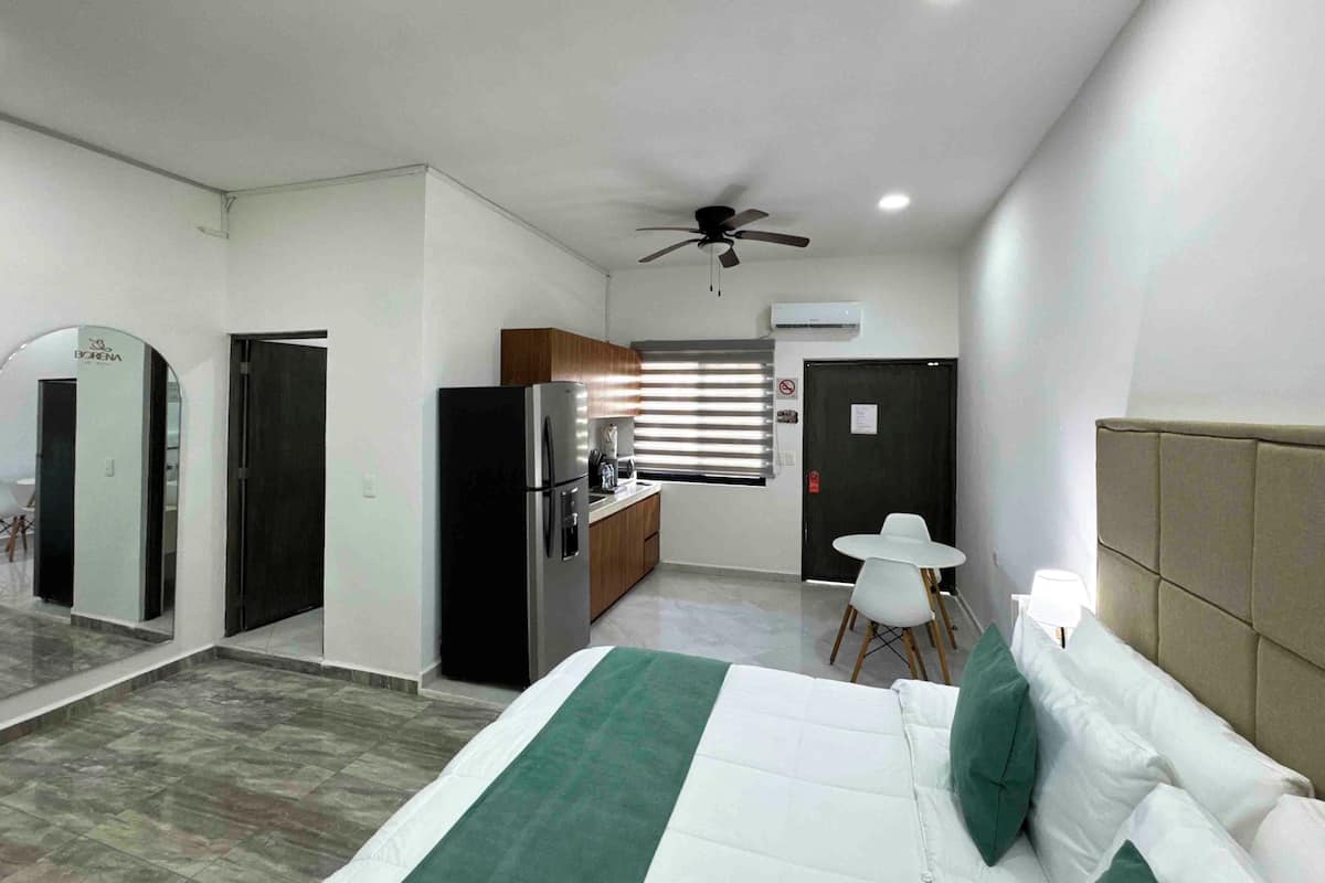 Quarto de hotel com cama, pequeno espaço de cozinha, ventoinha no teto, janela com persianas e espelho.