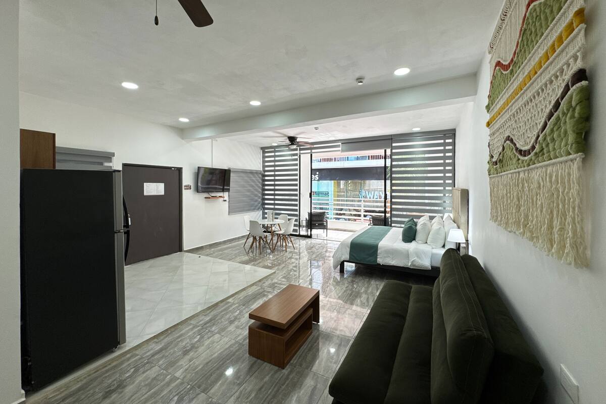 Quarto de hotel moderno com cama, sofá, televisão e área de jantar com cadeiras e mesa.