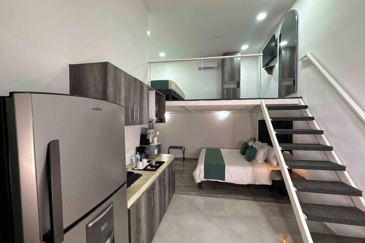 Cozinha compacta com geladeira, pia e bancada. Há uma cama com travesseiros e uma pequena mesa com cadeira. Uma escada leva a um andar superior.