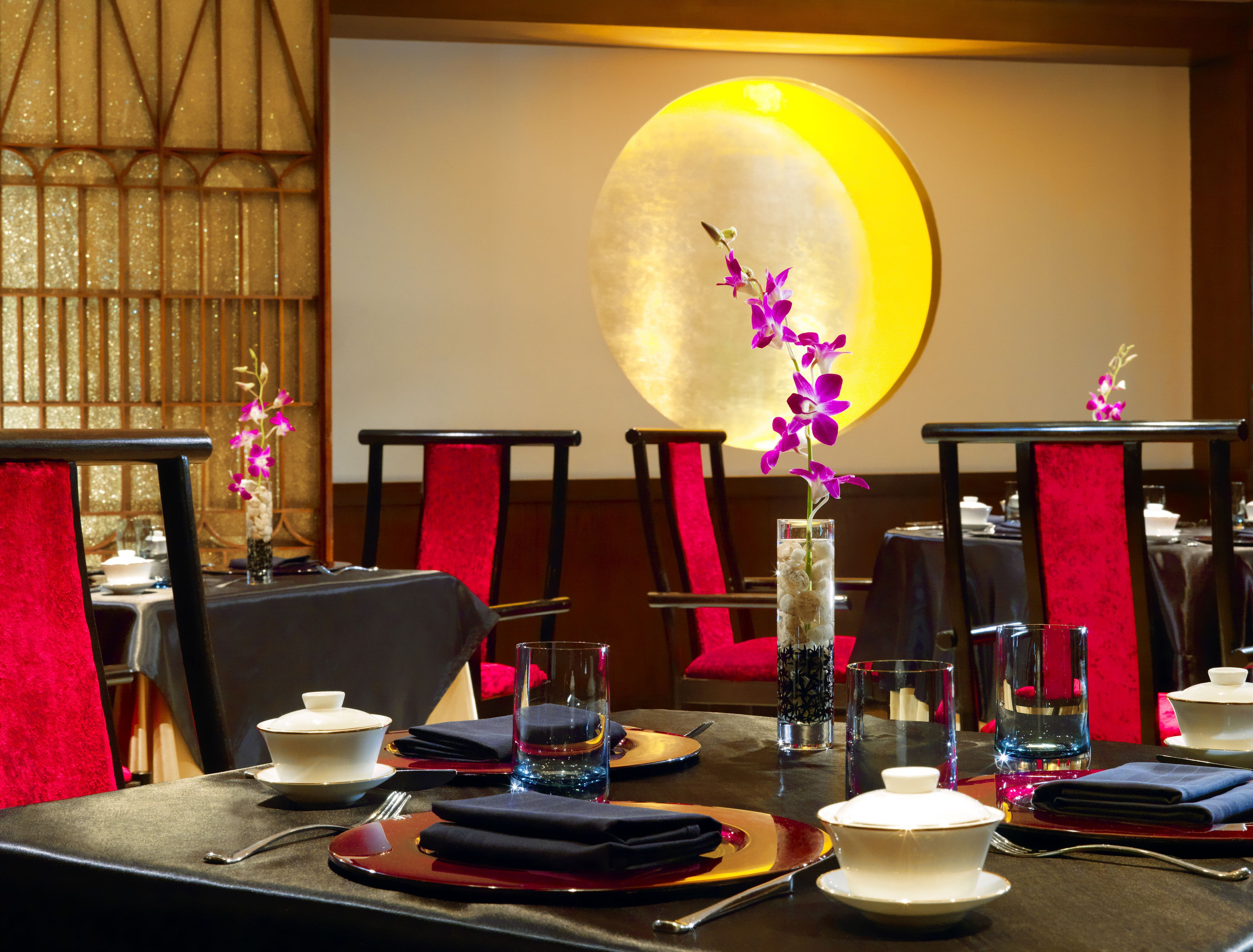 Uma sala de jantar com mesas preparadas para o serviço, com cadeiras vermelhas e pretas, louças brancas e orquídeas roxas em vasos.