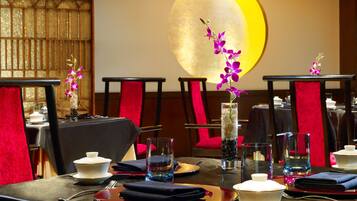 Uma sala de jantar com mesas preparadas para o serviço, com cadeiras vermelhas e pretas, louças brancas e orquídeas roxas em vasos.