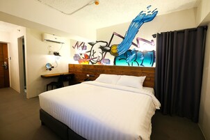 Quarto de hotel com cama, escrivaninha, aparelho de ar condicionado e parede com um mural colorido.