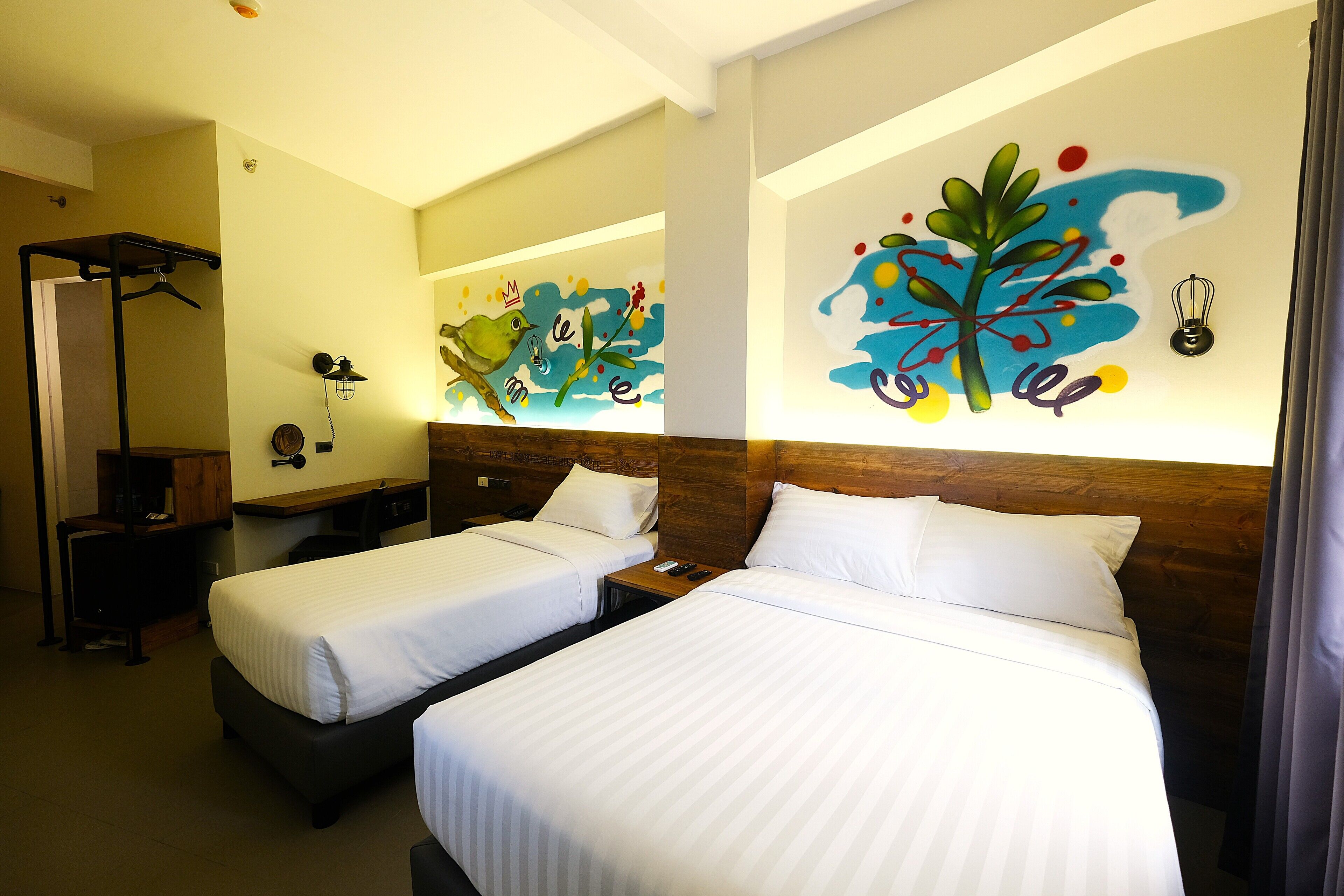 Quarto de hotel com duas camas, cabeceira de madeira, uma escrivaninha, uma cadeira, um cabideiro e um espelho.