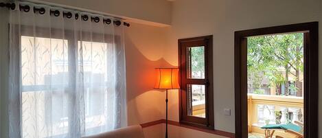 Quarto com ventilador de teto, sofá, luminária e vista para área externa com varanda.