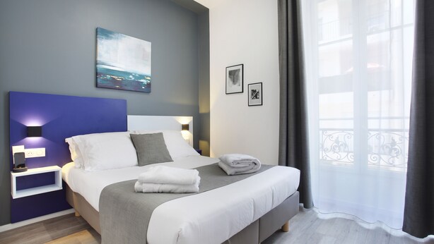 Quarto de hotel moderno com uma cama grande, uma mesa de cabeceira com abajur, um quadro pendurado na parede e uma janela com vista.