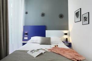 Quarto de hotel moderno com cabeceira azul, roupa de cama branca e cinzenta, manta rosa e uma revista na cama.