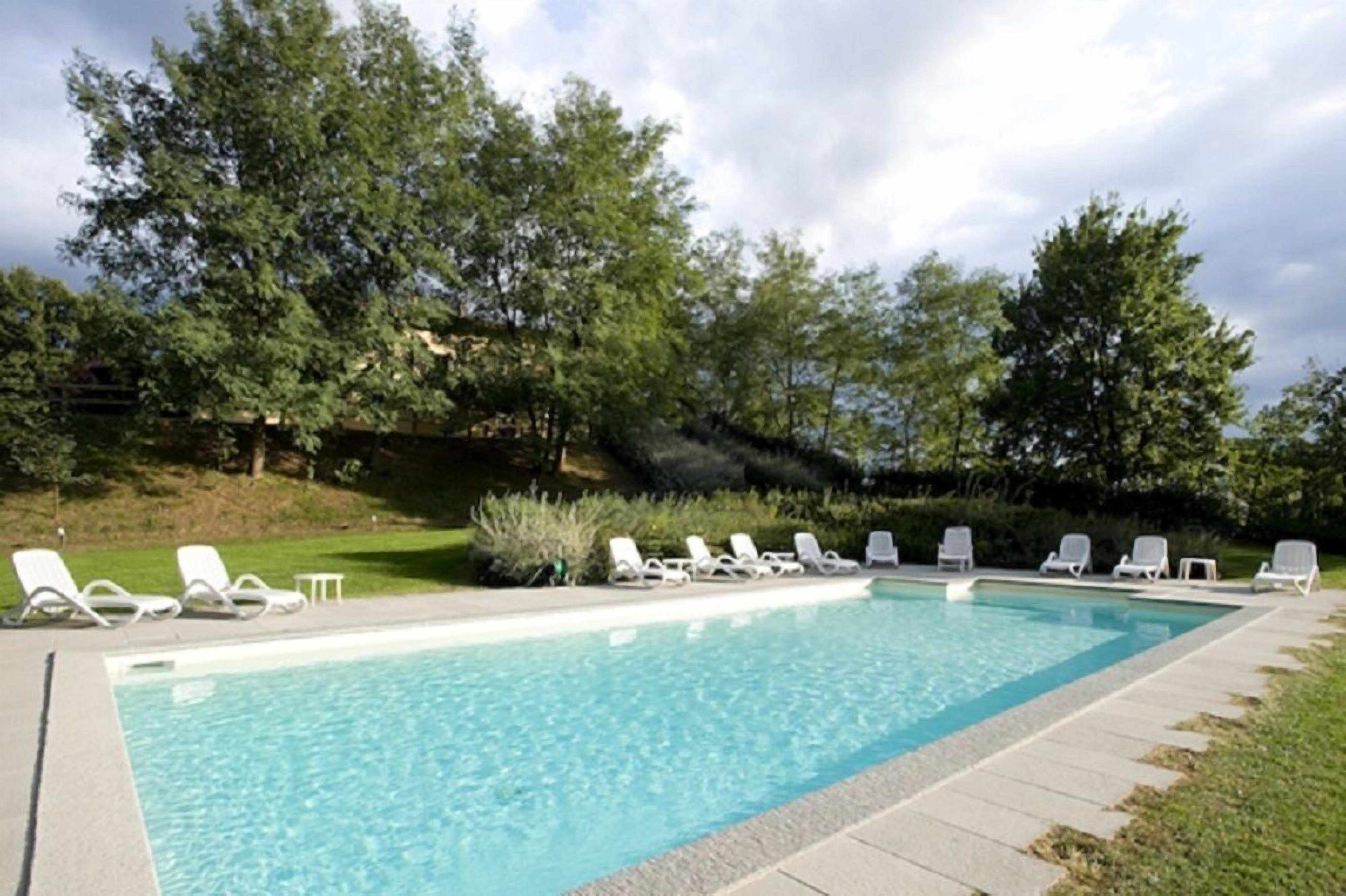 Piscina retangular com água azul cristalina, rodeada por espreguiçadeiras brancas e um gramado com árvores.