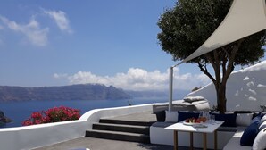 Uma área de estar ao ar livre com luz do sol, cadeiras de lounge brancas, uma mesa pequena e vista para o mar e montanhas.