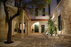 Um pátio com piso de pedra, uma árvore, plantas pendentes e um banco de metal.