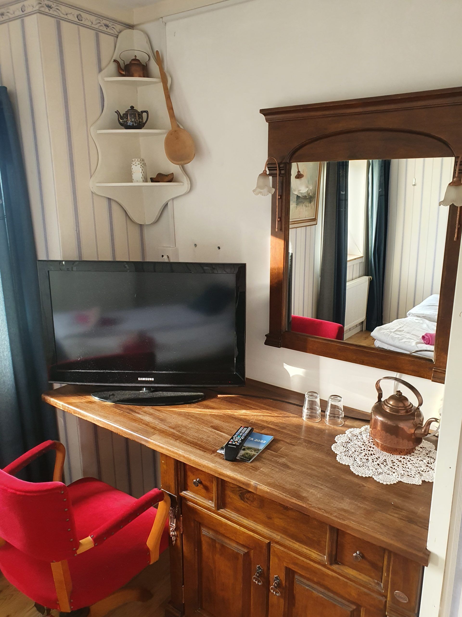 Quarto com televisão sobre suporte de madeira, uma cadeira vermelha, um armário de madeira com gavetas, uma estante com um bule de chá e um espelho refletindo uma janela com cortinas.