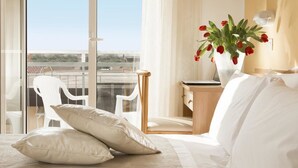 Quarto de hotel com cama, almofadas brancas, mesa de cabeceira com um vaso de tulipas vermelhas, um laptop e vista para o exterior através de grandes janelas.