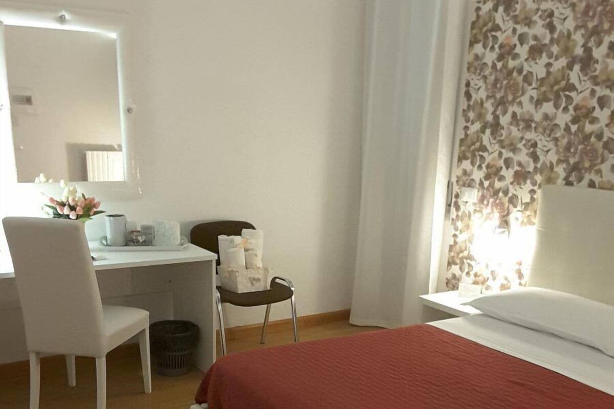 Quarto com cama, escrivaninha, cadeira, espelho e janela com cortinas.