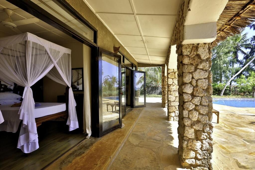 Quarto com cama sob uma rede mosquiteira, um pilar de pedra e vista para uma piscina.