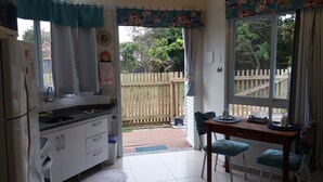 Uma cozinha compacta com geladeira, fogão e pia. Uma mesa de jantar com cadeiras está posicionada perto de uma janela grande, oferecendo vista para um deck de madeira e vegetação.