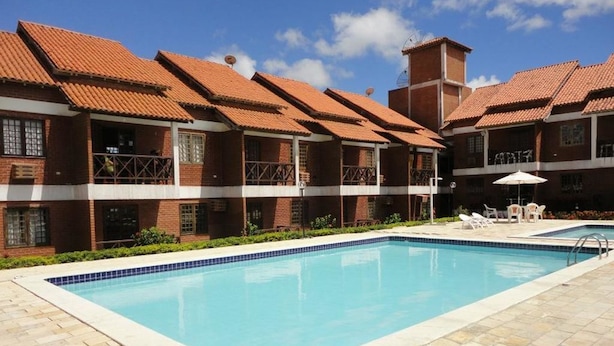 Uma piscina com deck revestido em azulejo, um edifício de tijolos com varandas e um céu azul límpido.